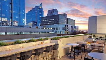 Um bar no terraço com mesas ao ar livre, o horizonte da cidade ao fundo e um céu ao pôr do sol.