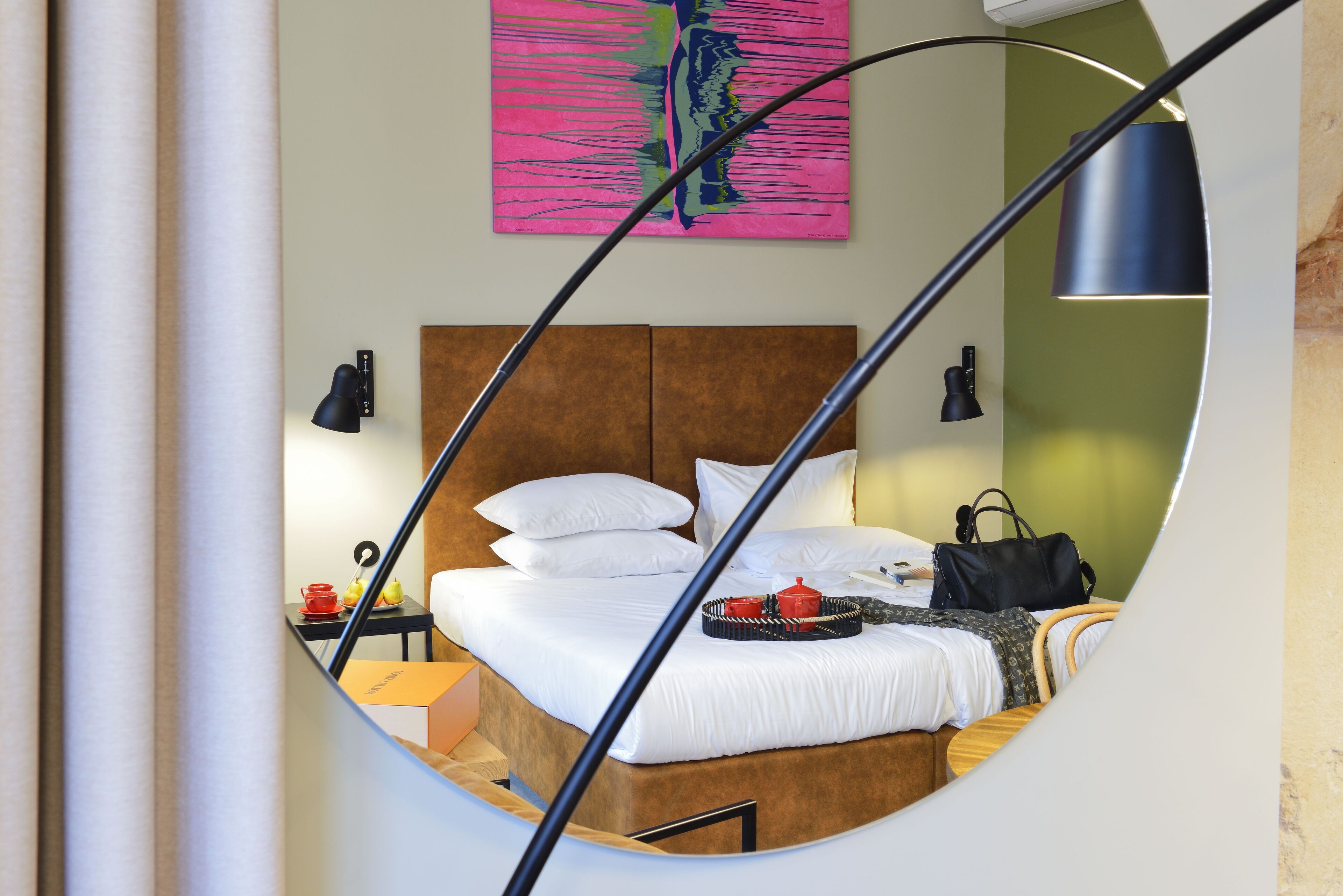 Um espelho redondo reflete um quarto moderno com cabeceira de madeira, roupa de cama branca, uma chaleira vermelha e uma bolsa preta.