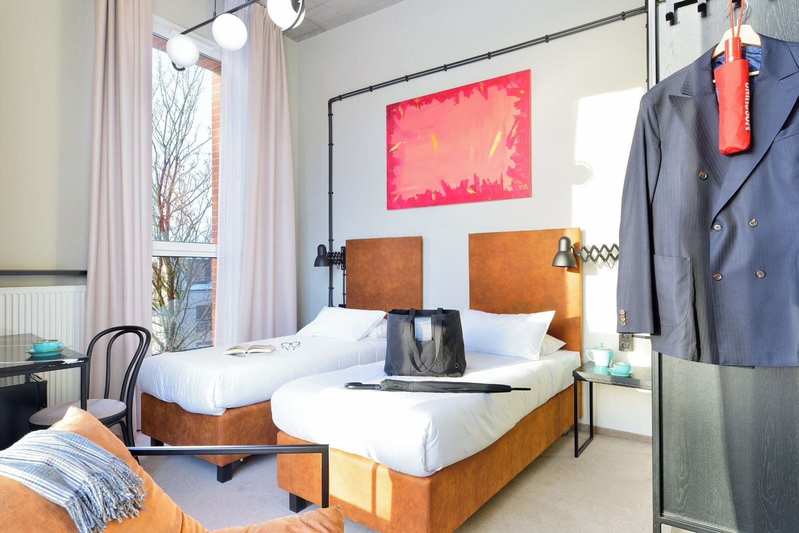 Quarto de hotel com duas camas, janela com cortinas, um quadro abstrato vermelho, um cabideiro com um casaco e uma mesa com abajur.