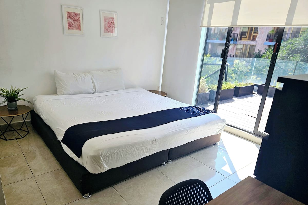 Quarto moderno com uma cama grande, uma mesinha com uma planta e vista para a varanda.