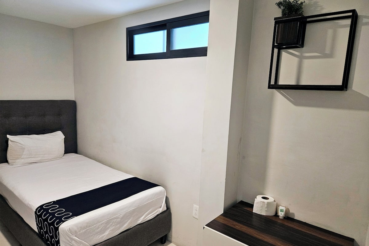 Um quarto com uma cama, uma janela pequena, uma mesa de cabeceira com uma planta e uma prateleira na parede.