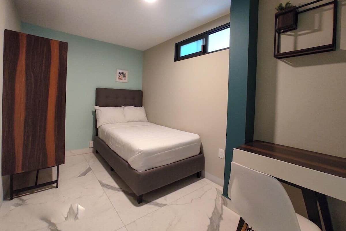 Um quarto com uma cama, penteadeira, janela pequena e prateleira fixada na parede.