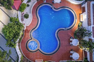 Vista aérea de uma piscina com design de borda ondulada, rodeada por um deck de ladrilhos vermelhos e mobiliário de área externa com guarda-sóis.