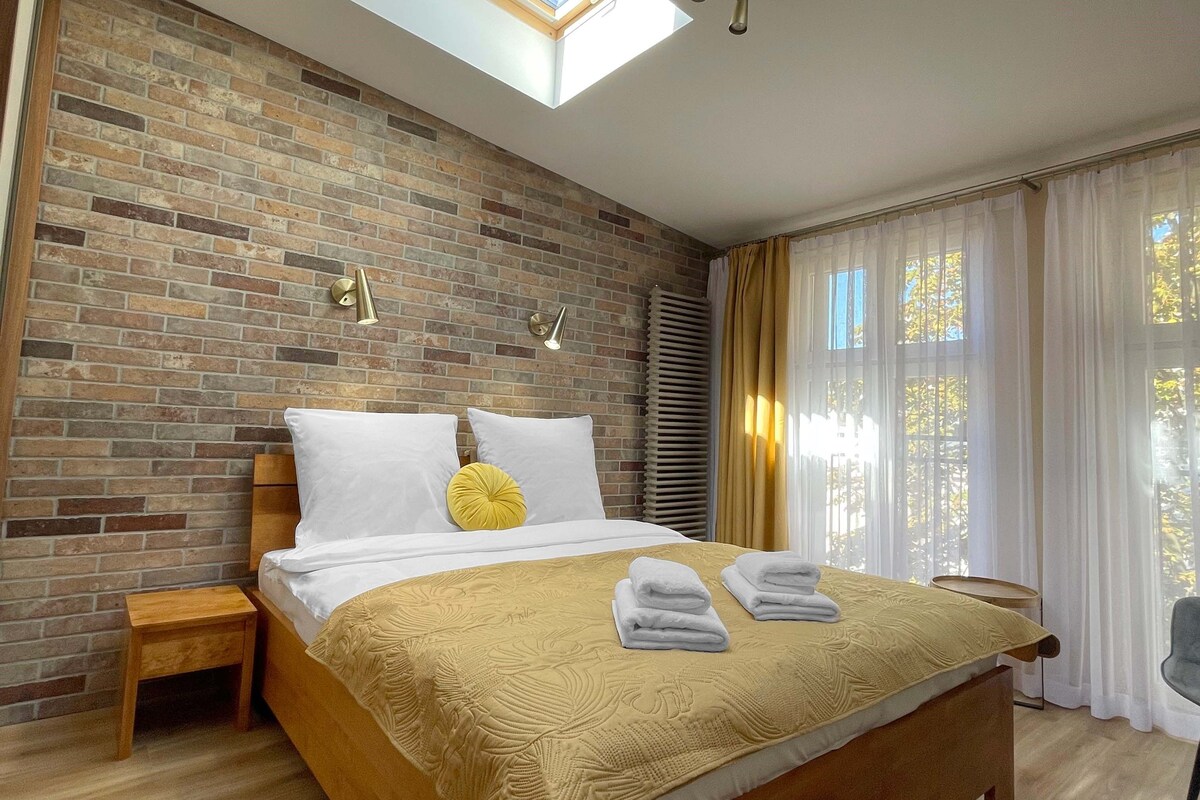 Quarto moderno com claraboia, parede de tijolos e cama com roupa de cama branca e travesseiro amarelo.
