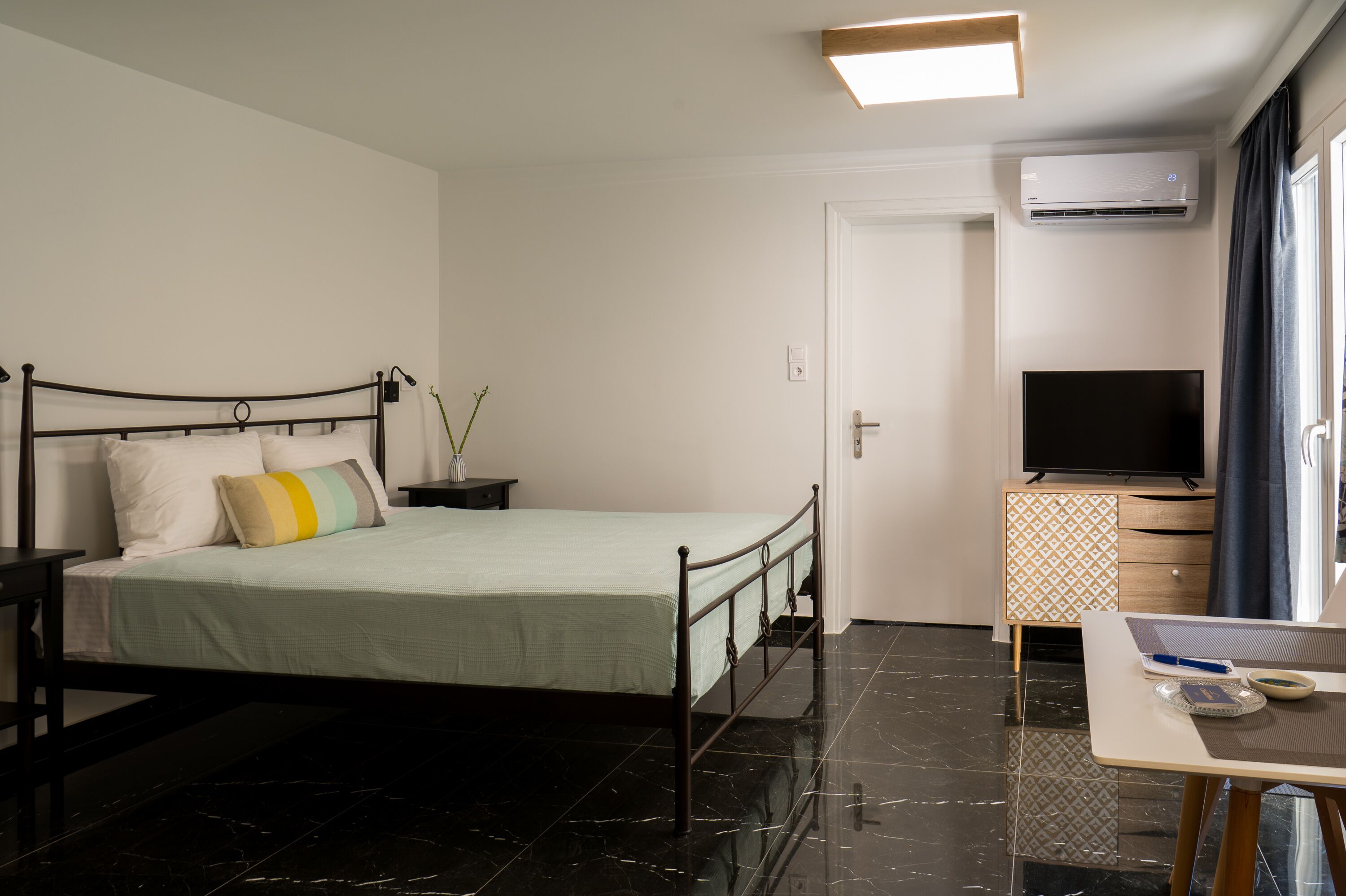 Quarto de hotel moderno com cama, secretária e televisão, e porta de acesso ao banheiro.