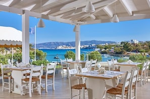 Restaurante à beira-mar com mesas ao ar livre, mobiliário branco e vista para o mar e edifícios.