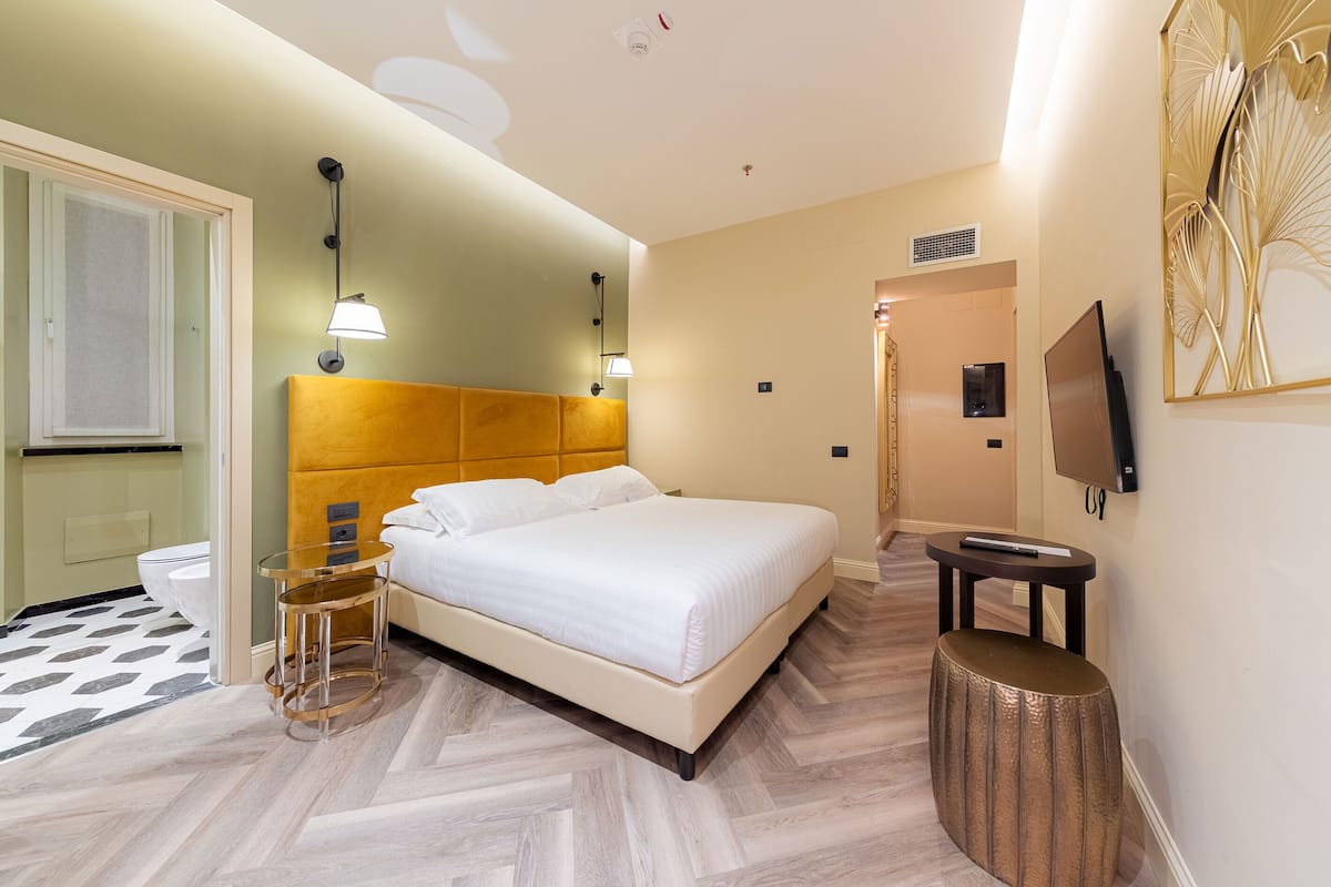 Quarto de hotel com cama, mesa de cabeceira, luminária, televisão e banheiro com vaso sanitário e chuveiro.