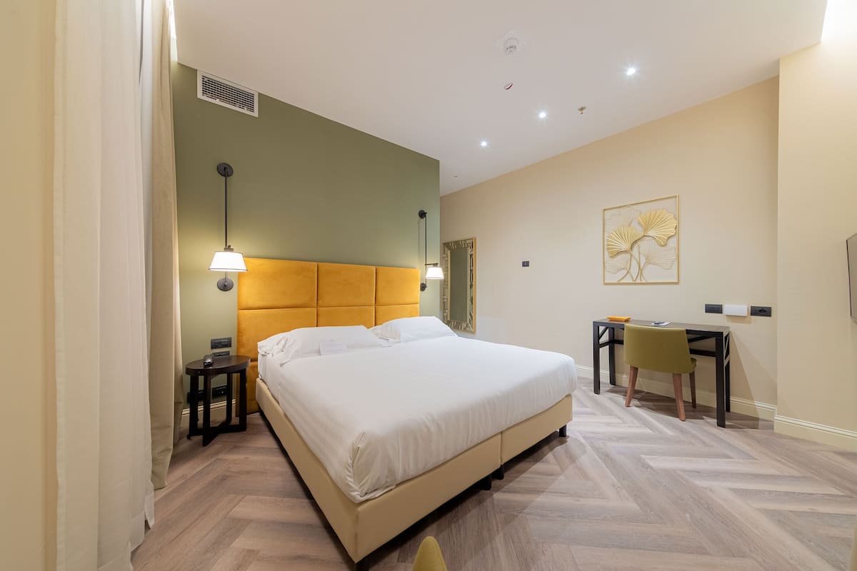 Quarto de hotel com cama, mesa de cabeceira, luminária, escrivaninha, cadeira e obra de arte na parede.