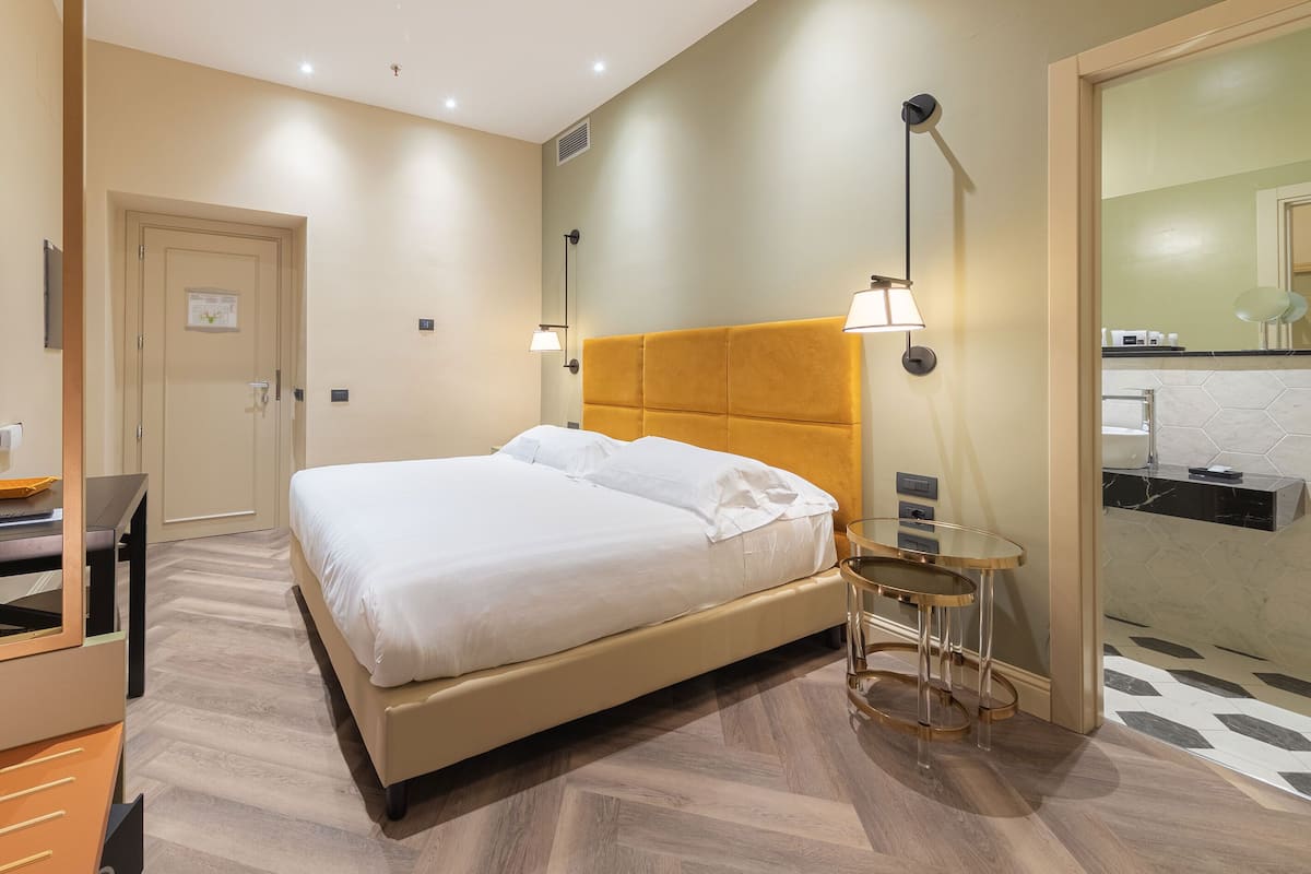 Quarto de hotel com uma cama, mesa de cabeceira, abajur e um banheiro visível por uma porta aberta.