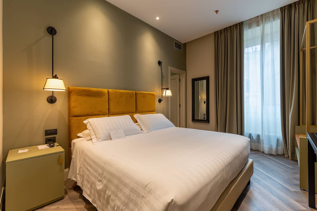 Uma cama bem arrumada com lençóis brancos, uma mesa de cabeceira com abajur e uma janela grande com cortinas.