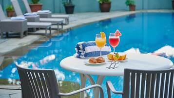 Mesa à beira da piscina com bebidas e petiscos, rodeada por espreguiçadeiras e plantas em vasos.