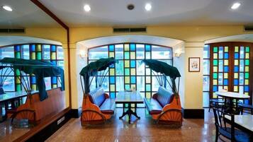 Restaurante com área de estar central, com bancos estofados e mesas, rodeado por grandes janelas com painéis de vidro coloridos.