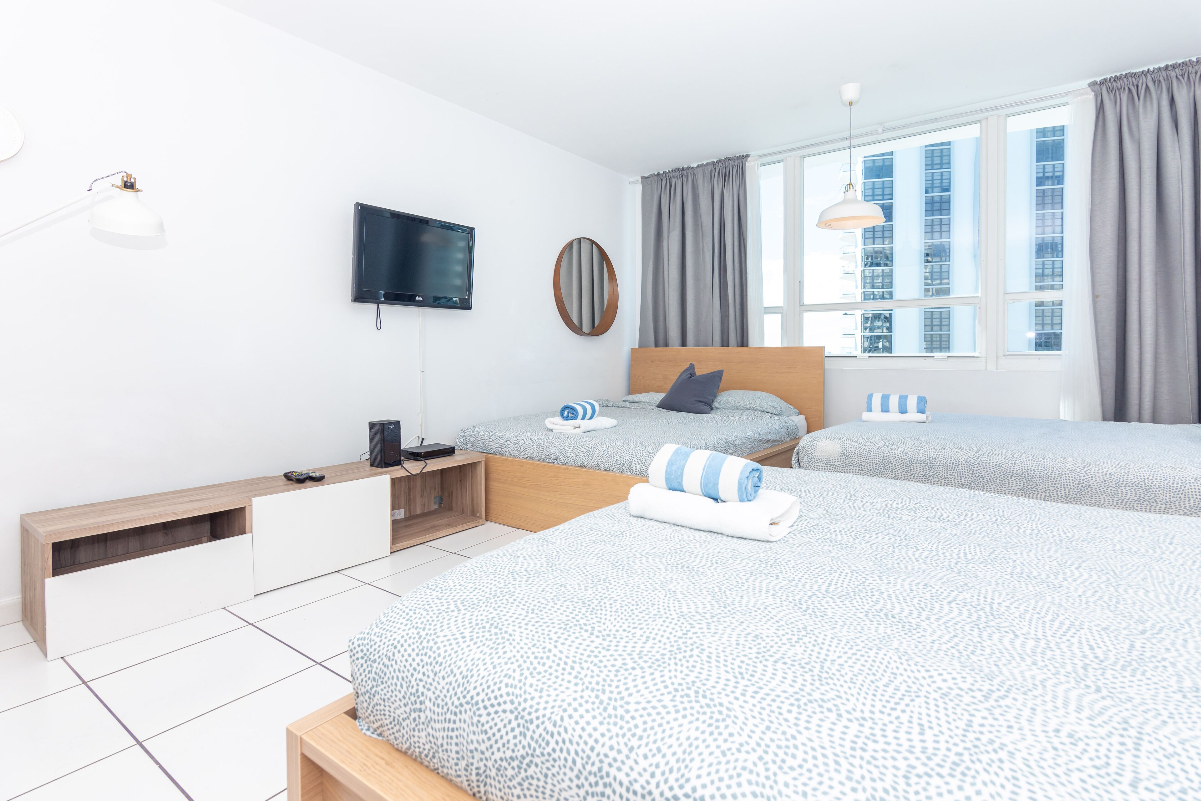 Quarto de hotel moderno com duas camas, televisão, escrivaninha e uma janela grande com cortinas.