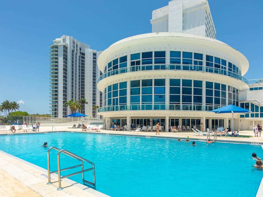 Área de piscina com um prédio circular grande, um prédio alto adjacente e céu claro.