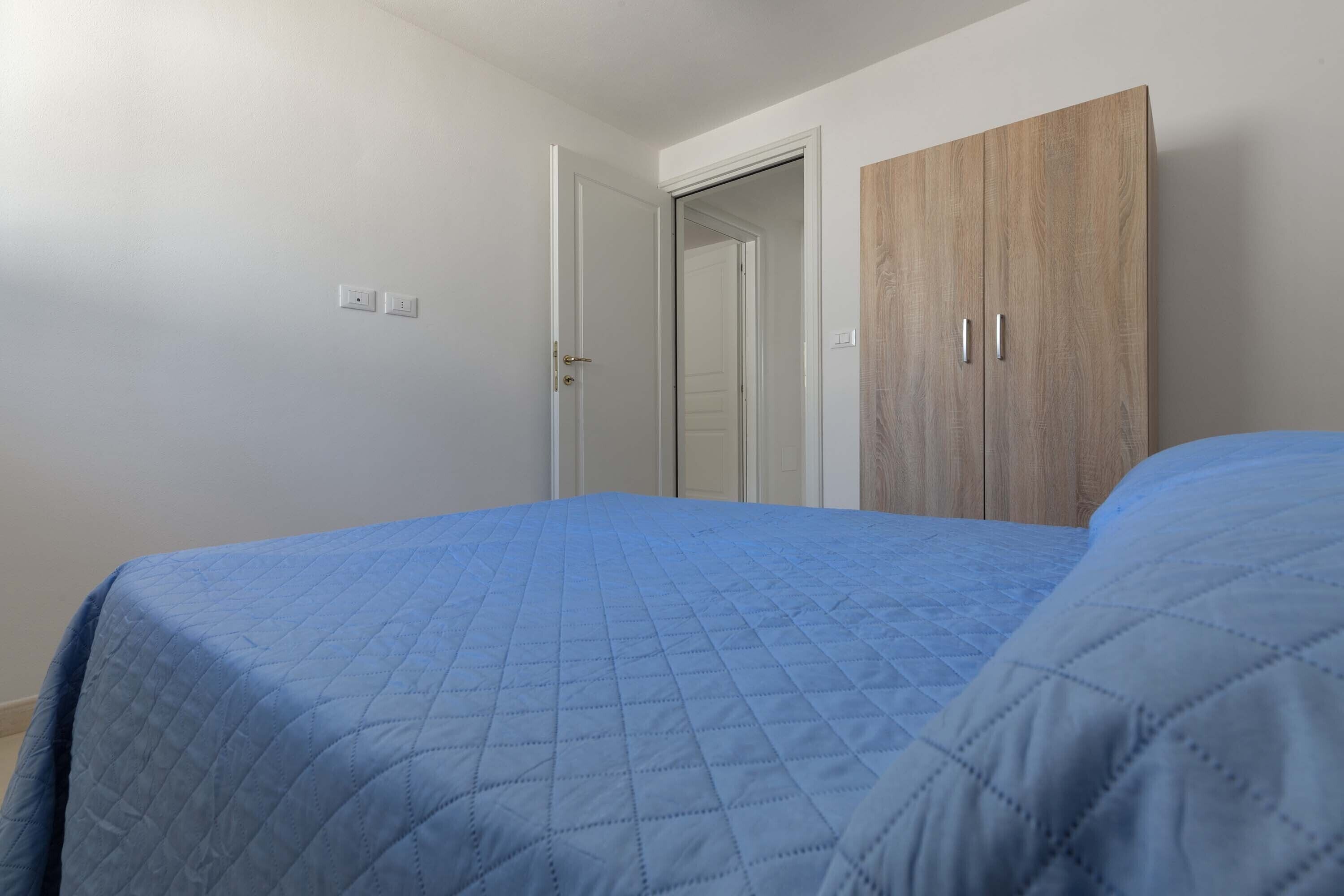 Quarto com colcha azul acolchoada, guarda-roupa de madeira e porta branca.