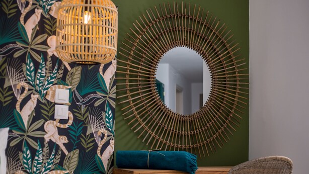 Um quarto com parede verde, luminária trançada, espelho circular, cômoda de madeira e cadeira de vime.