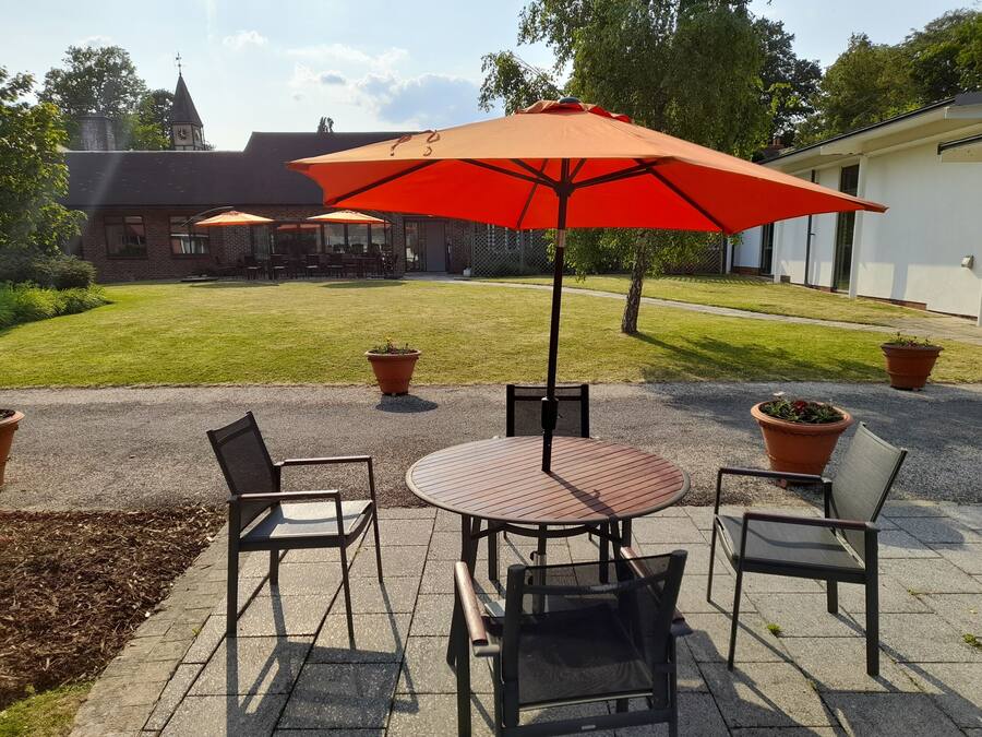 Uma área de pátio com uma mesa redonda e quatro cadeiras, um grande guarda-sol vermelho e plantas em vasos.