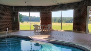 Piscina com deck revestido, uma pequena mesa com cadeiras e grandes janelas com vista para um campo gramado.