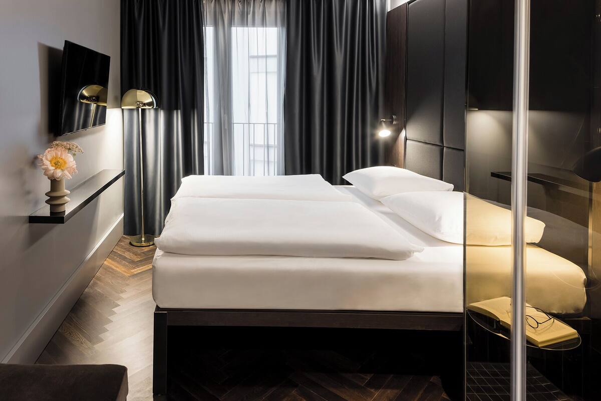 Quarto de hotel moderno com uma cama grande, mesa de cabeceira, televisão e janela com cortinas.
