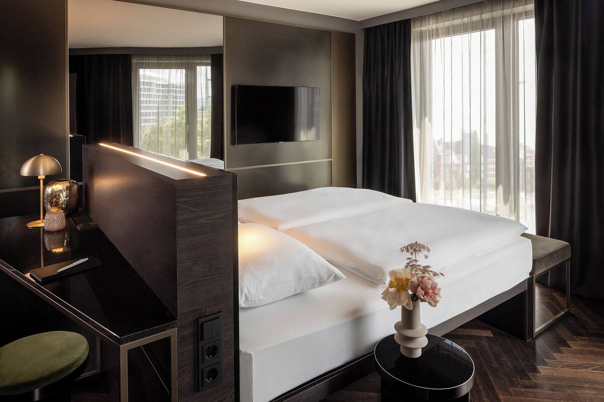 Quarto de hotel moderno com uma cama grande, uma escrivaninha com luminária, uma TV de tela plana na parede e vista da cidade através de grandes janelas.