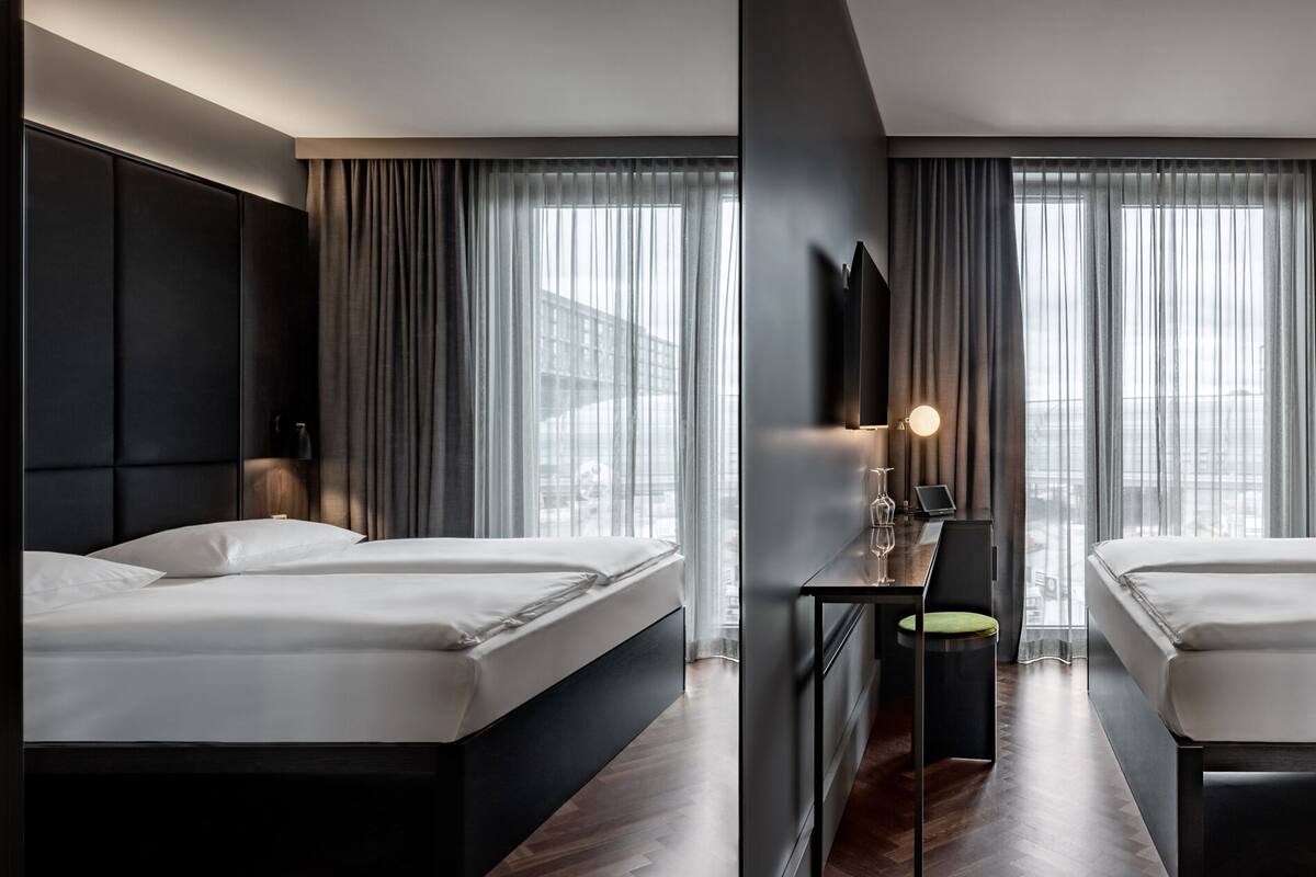 Quarto de hotel moderno com duas camas, uma janela grande com cortinas, uma TV de tela plana na parede e uma mesinha com luminária.