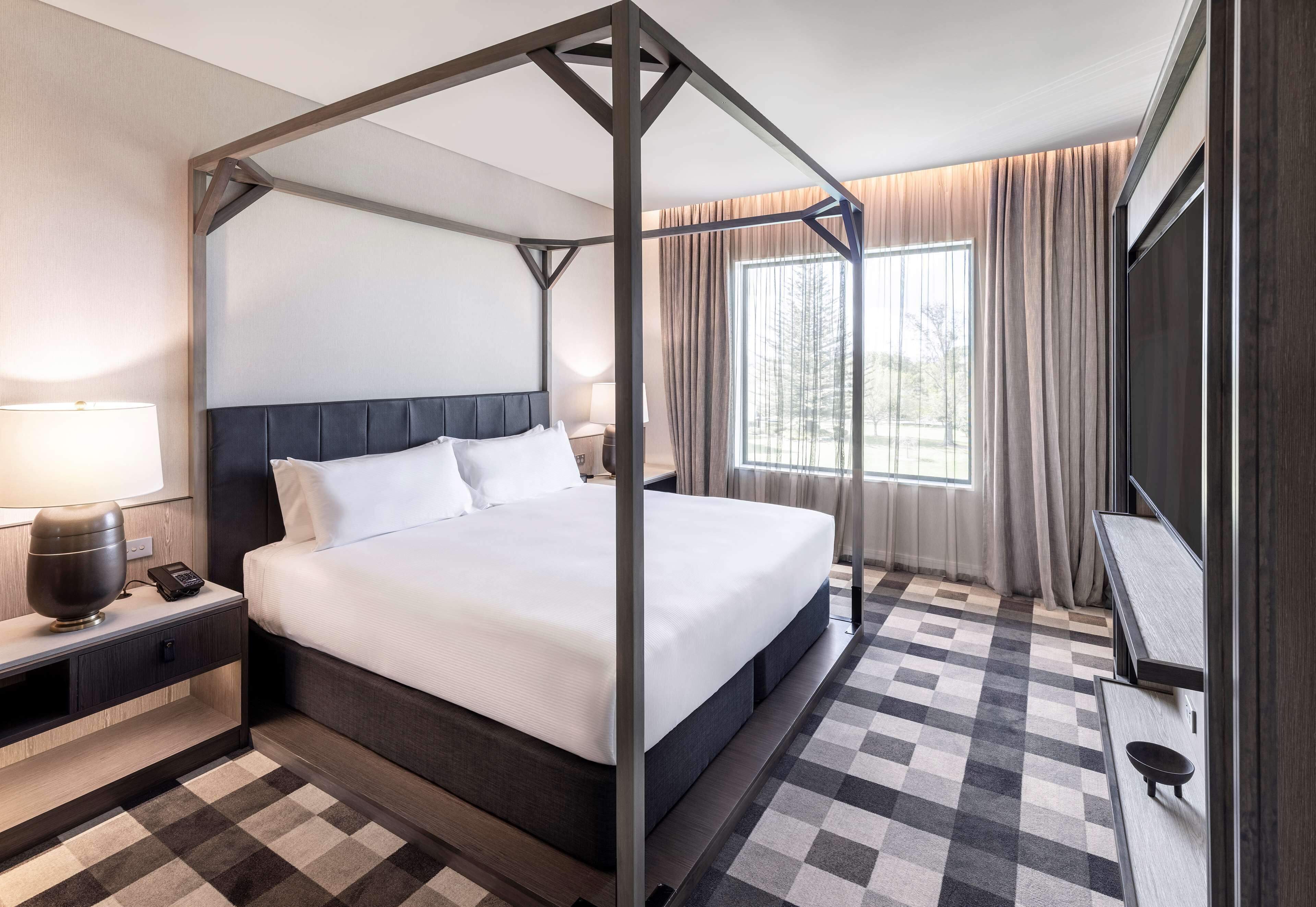 Quarto de hotel moderno com uma cama de dossel, uma escrivaninha com luminária e uma grande janela com cortinas.