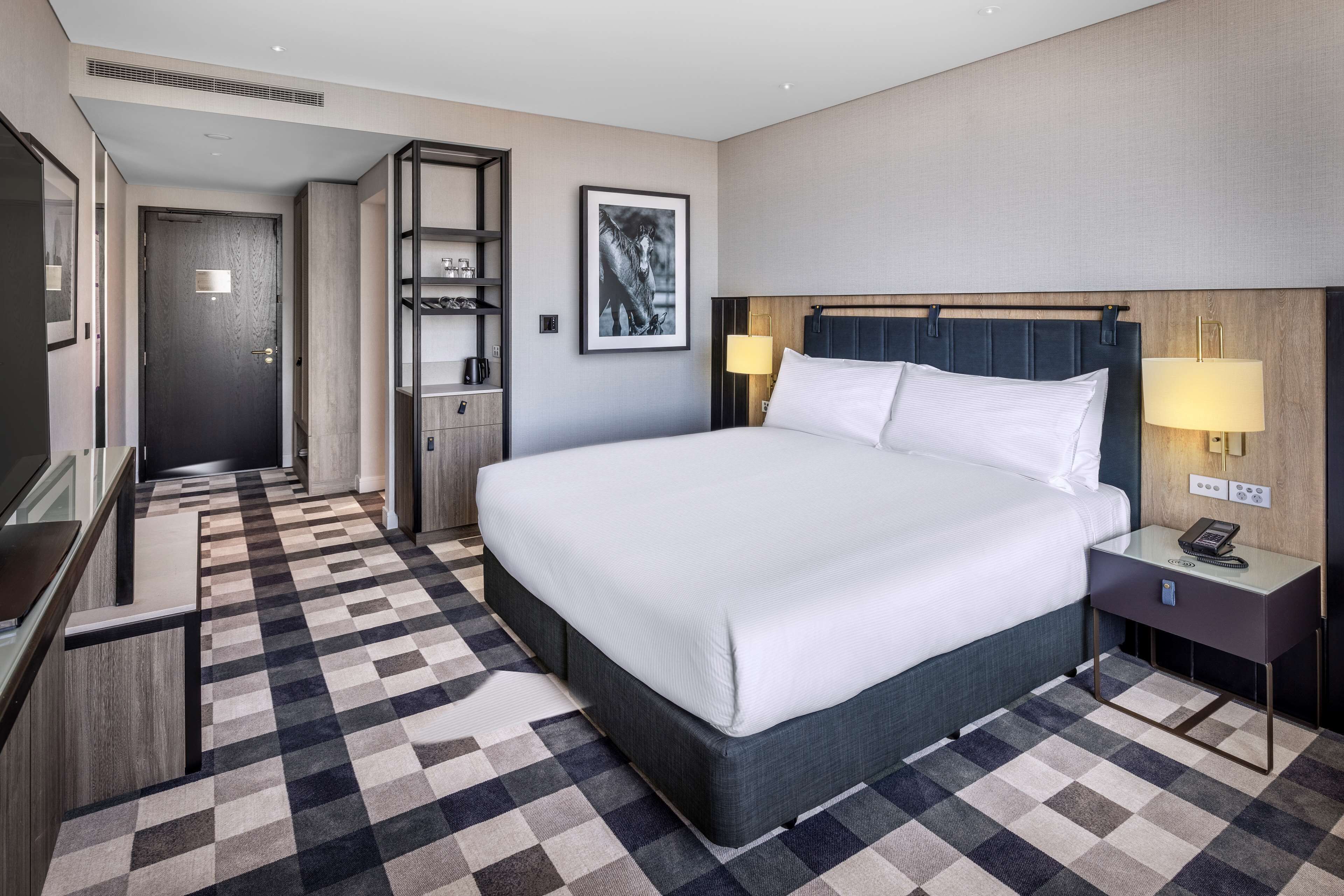 Quarto de hotel com uma cama grande, duas mesas de cabeceira com luminárias, um guarda-roupa, uma escrivaninha e uma televisão.