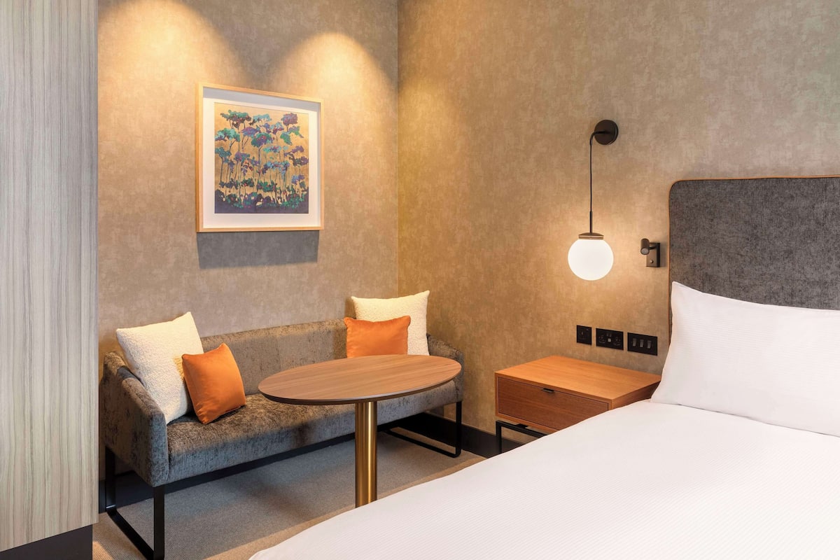 Quarto de hotel moderno com um sofá cinza, uma mesa redonda de madeira, uma cama de solteiro e um candeeiro fixado na parede.