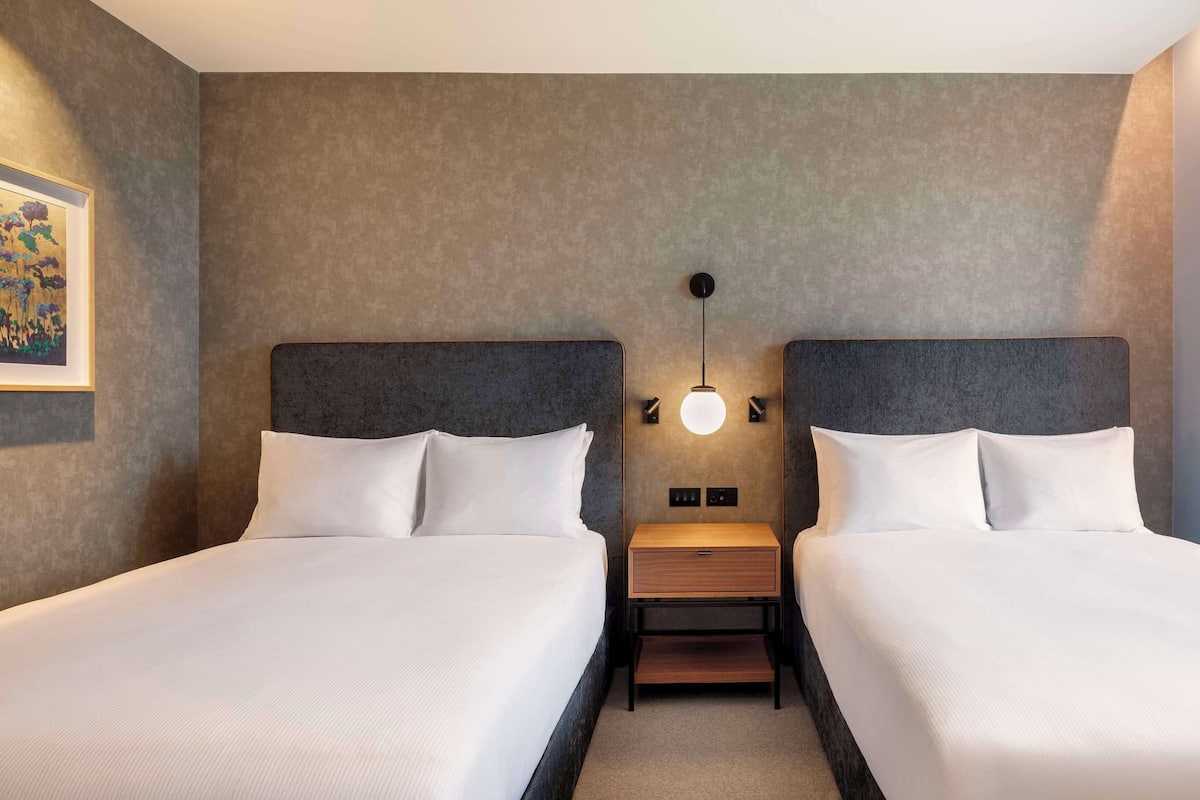 Quarto de hotel com duas camas, uma mesinha de cabeceira, uma luminária na parede e um quadro emoldurado.