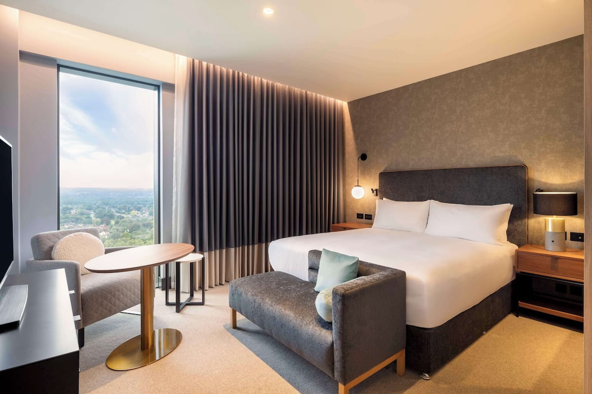 Quarto de hotel moderno com uma cama grande, uma escrivaninha, uma cadeira e vista para a cidade.