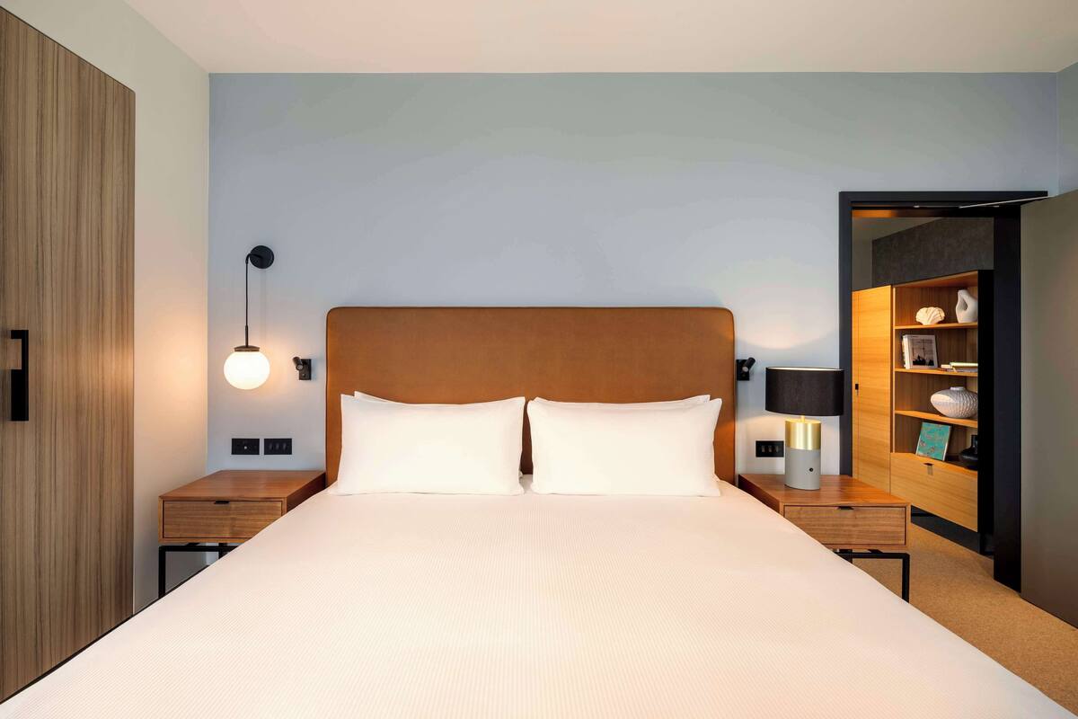 Quarto de hotel com uma cama grande, cabeceira de madeira, mesas de cabeceira, uma luminária e um criado-mudo.
