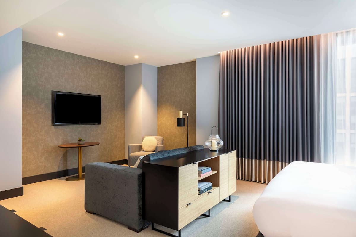 Quarto de hotel moderno com televisão de tela plana na parede, um sofá, uma mesinha de cabeceira com abajur, uma cama e uma janela grande com cortinas.