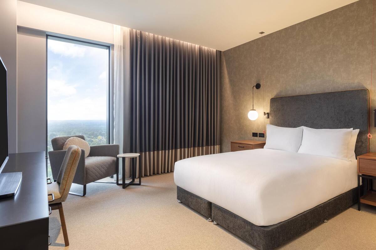 Quarto de hotel moderno com uma cama grande, uma escrivaninha, uma cadeira e vista para a cidade.