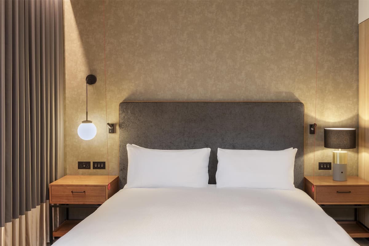 Quarto de hotel moderno com uma cama grande, duas mesas de cabeceira, encosto de cama e iluminação embutida na parede.