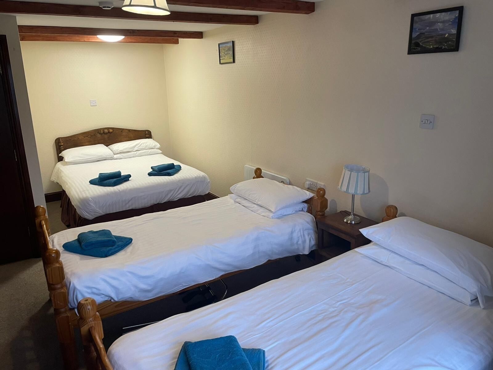 Quarto de hotel com três camas de solteiro, cabeceiras de madeira, roupa de cama branca e uma luminária de cabeceira.