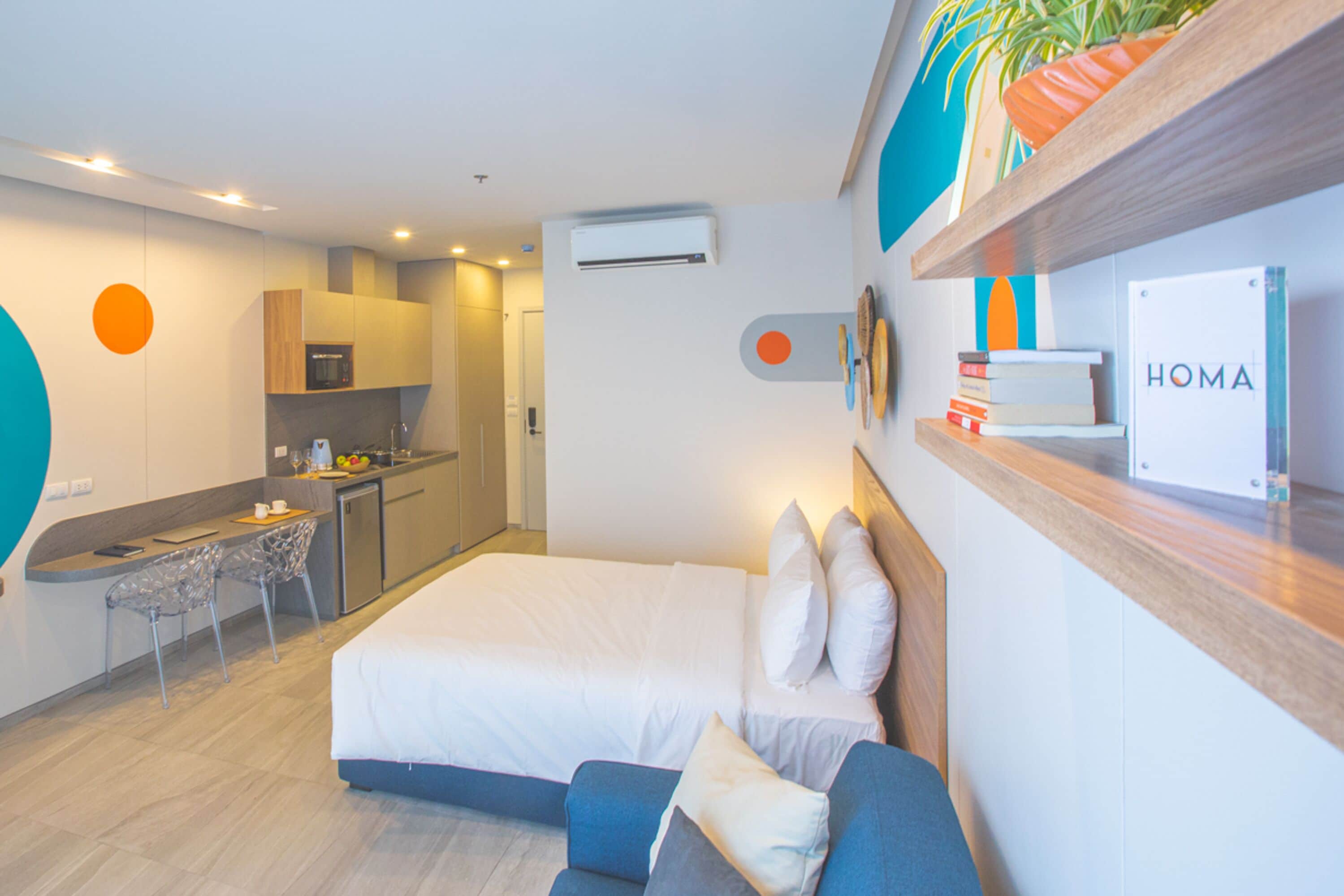 Quarto de hotel moderno com cama, sofá, escrivaninha e cozinha compacta.