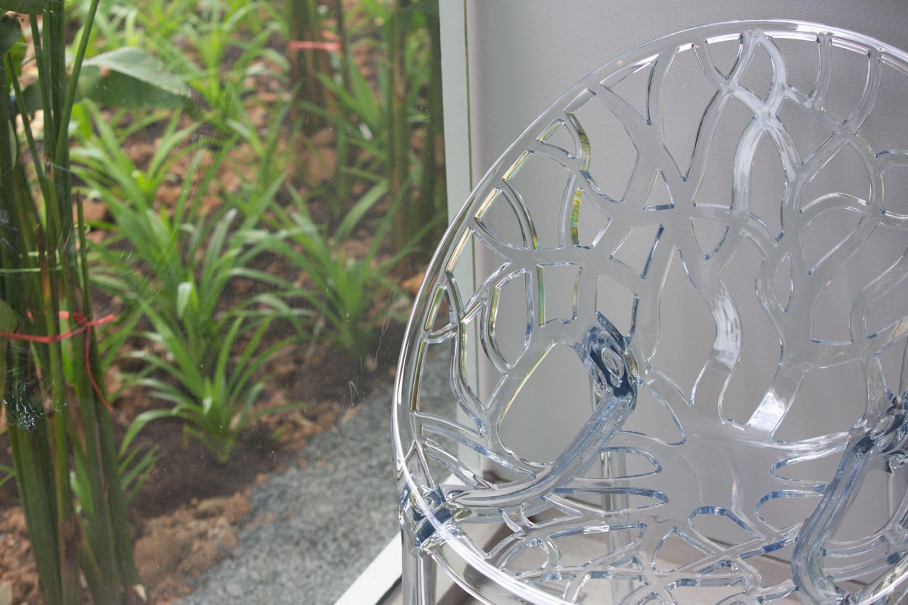Uma mesa de vidro transparente com padrão geométrico, colocada ao lado de uma janela com vista para plantas verdes.