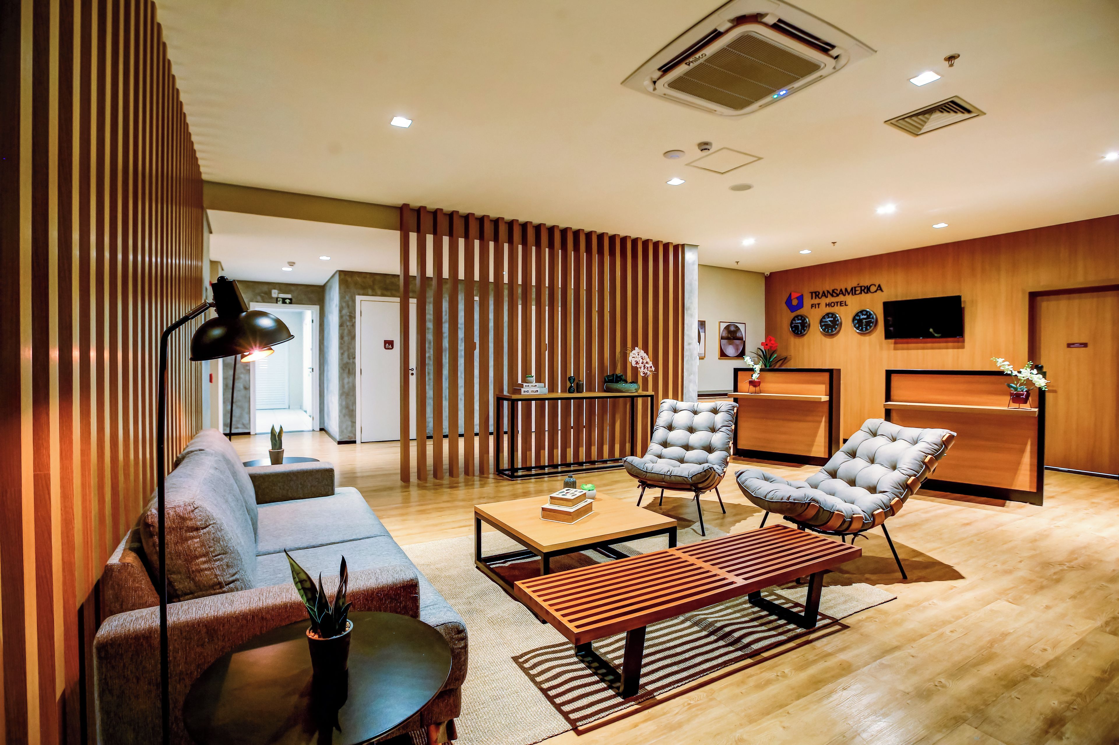 Um hall de hotel moderno com um balcão de recepção de madeira, um sofá, duas poltronas, uma mesa de centro e uma luminária.