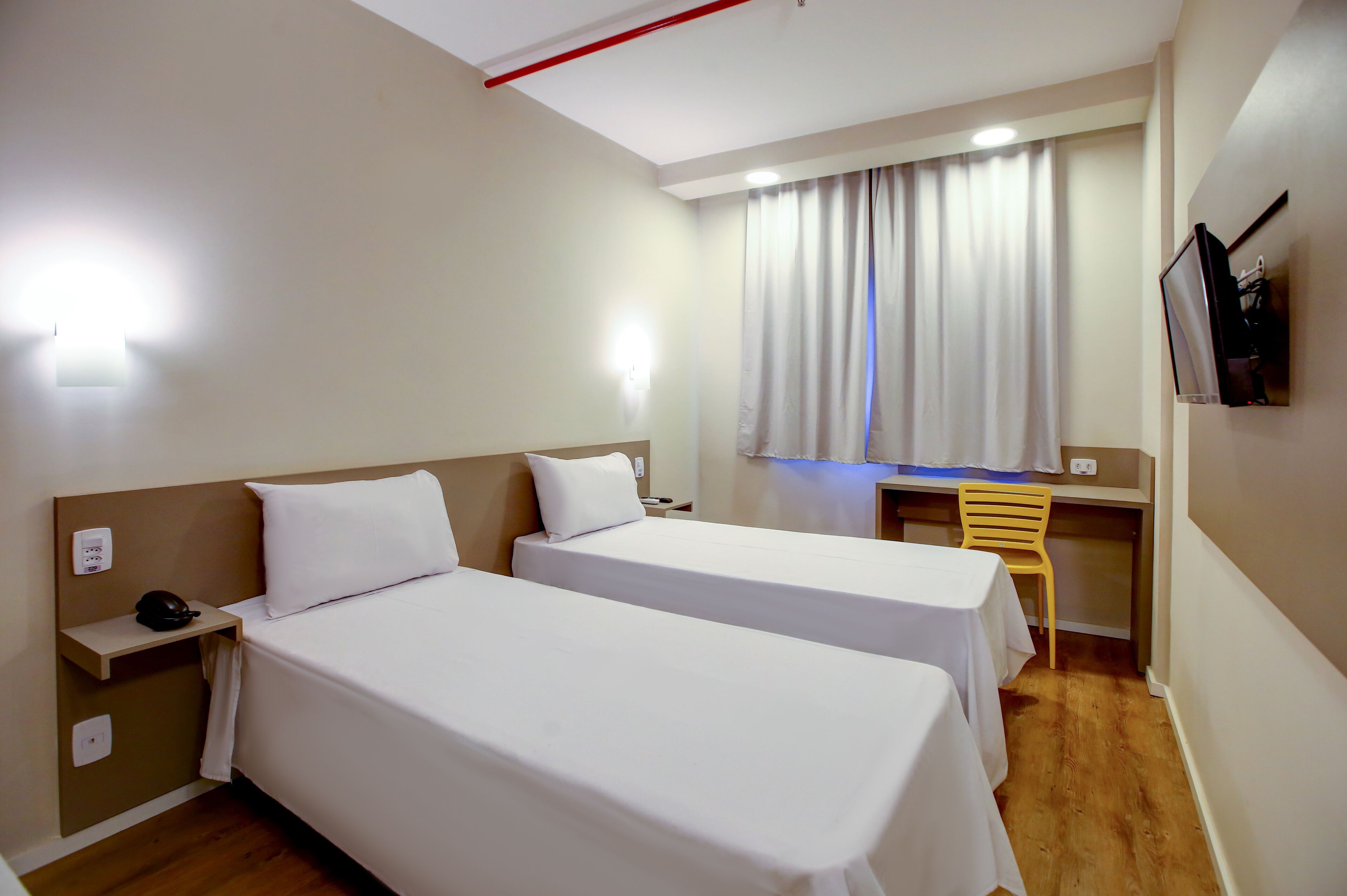 Quarto de hotel com duas camas de solteiro, uma escrivaninha, uma cadeira, televisão fixada na parede e uma janela com cortinas.