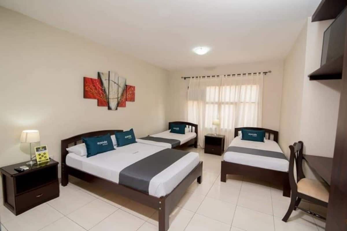 Quarto de hotel com duas camas de solteiro, uma escrivaninha, uma cadeira, mesa de cabeceira com abajur e TV fixada na parede.