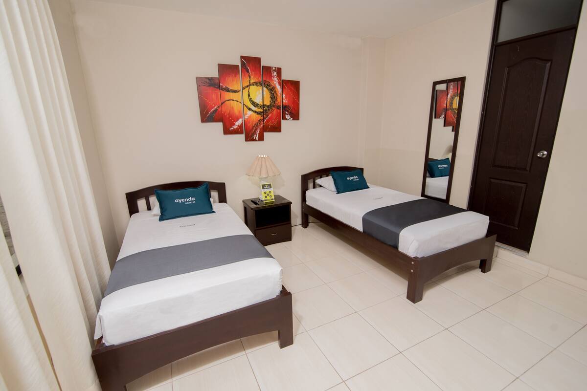 Quarto de hotel com duas camas, mesa de cabeceira, luminária, espelho e um quadro na parede.
