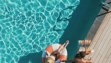 Uma piscina com água cristalina, uma mulher de fato de banho e chapéu de palha flutuando em um círculo inflável laranja e branco, e um homem parado na beira da piscina.