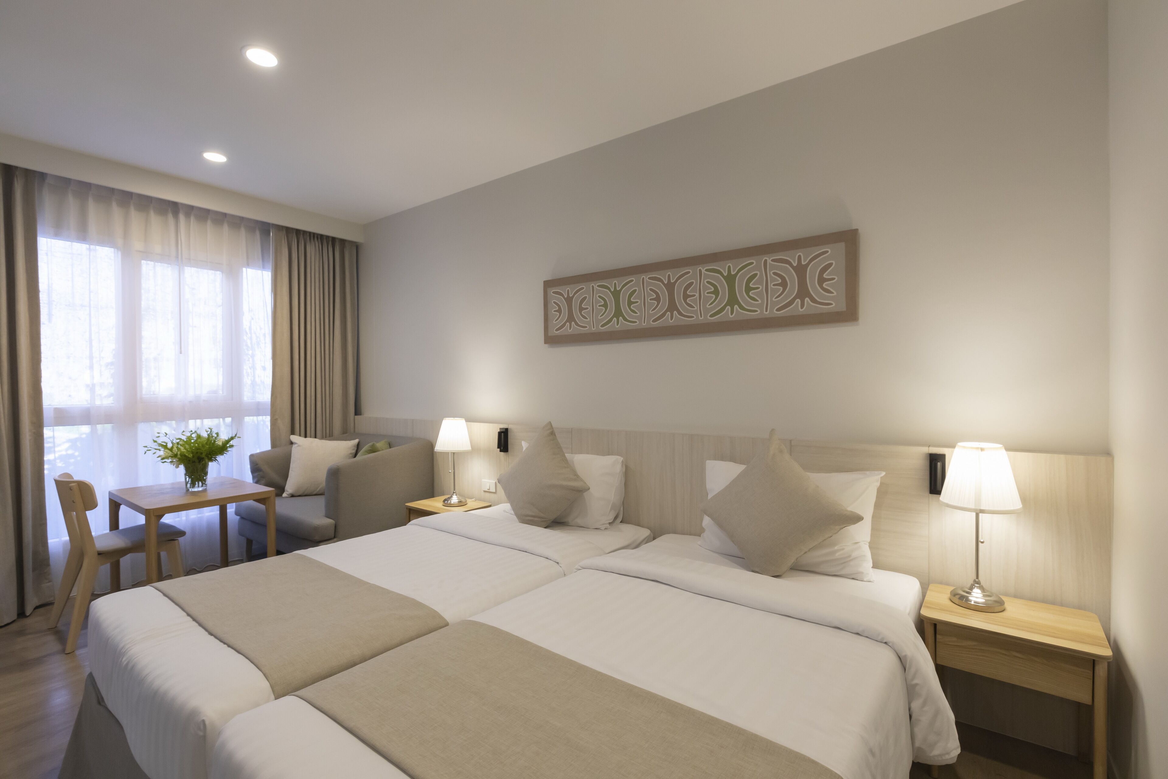Quarto de hotel com uma cama grande, uma secretária e uma cadeira. Há uma janela com cortinas, uma obra de arte na parede e uma mesa de cabeceira com um abajur.