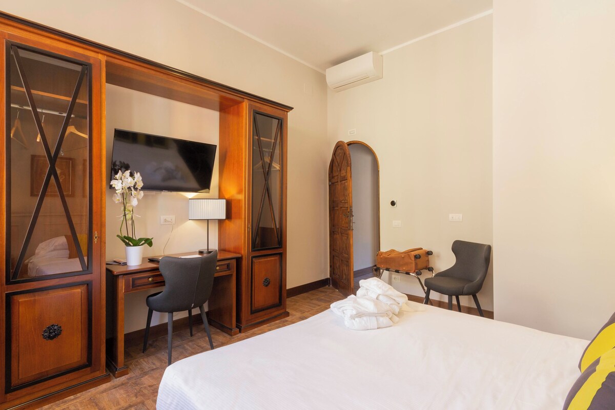 Quarto de hotel com cama, escrivaninha, cadeira, televisão, guarda-roupa e espelho.