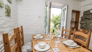 Uma sala de jantar com uma mesa de madeira posta para quatro pessoas, cercada por cadeiras de madeira. Uma janela oferece vista para o verde lá fora.