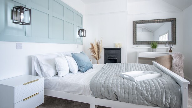 Quarto com cama branca, mesa de cabeceira, cômoda, espelho e luminária fixa na parede.