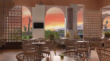Um restaurante com cadeiras e mesas de vime, vista para o pôr do sol através de aberturas em arco e um painel decorativo.