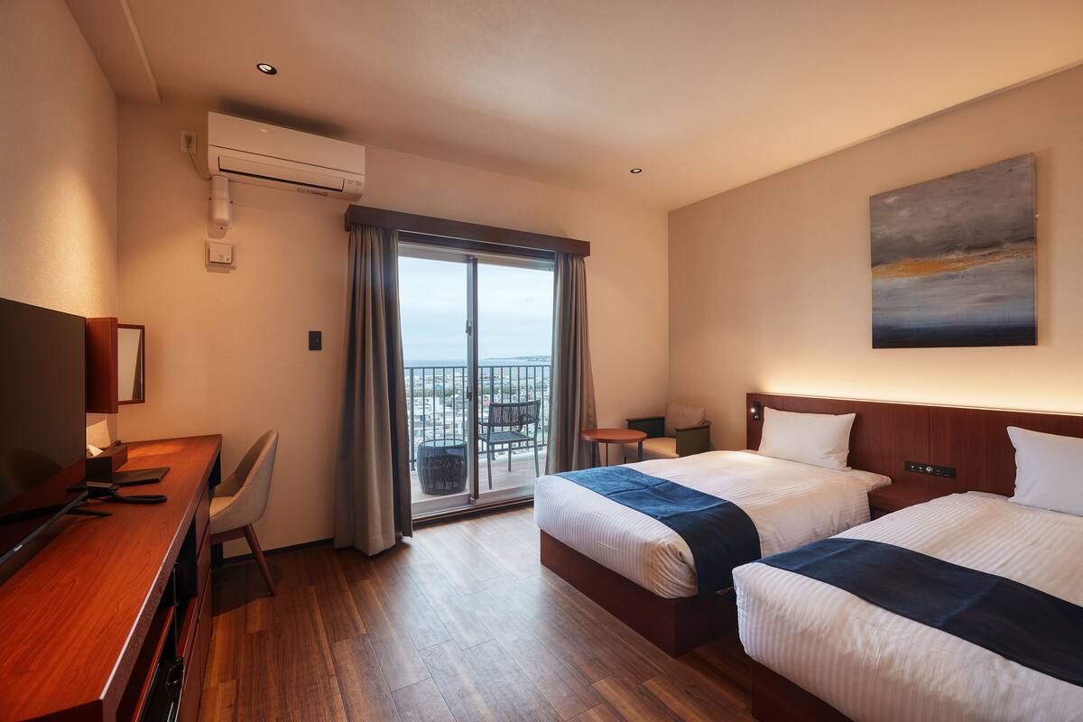 Quarto de hotel com duas camas, uma escrivaninha, uma cadeira, uma televisão e varanda com vista.