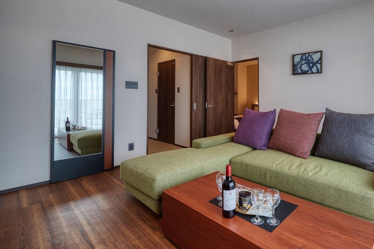 Quarto de hotel moderno com um sofá verde, uma mesa de centro de madeira com uma garrafa e copos, uma cama com edredom azul e branco, e um espelho grande.