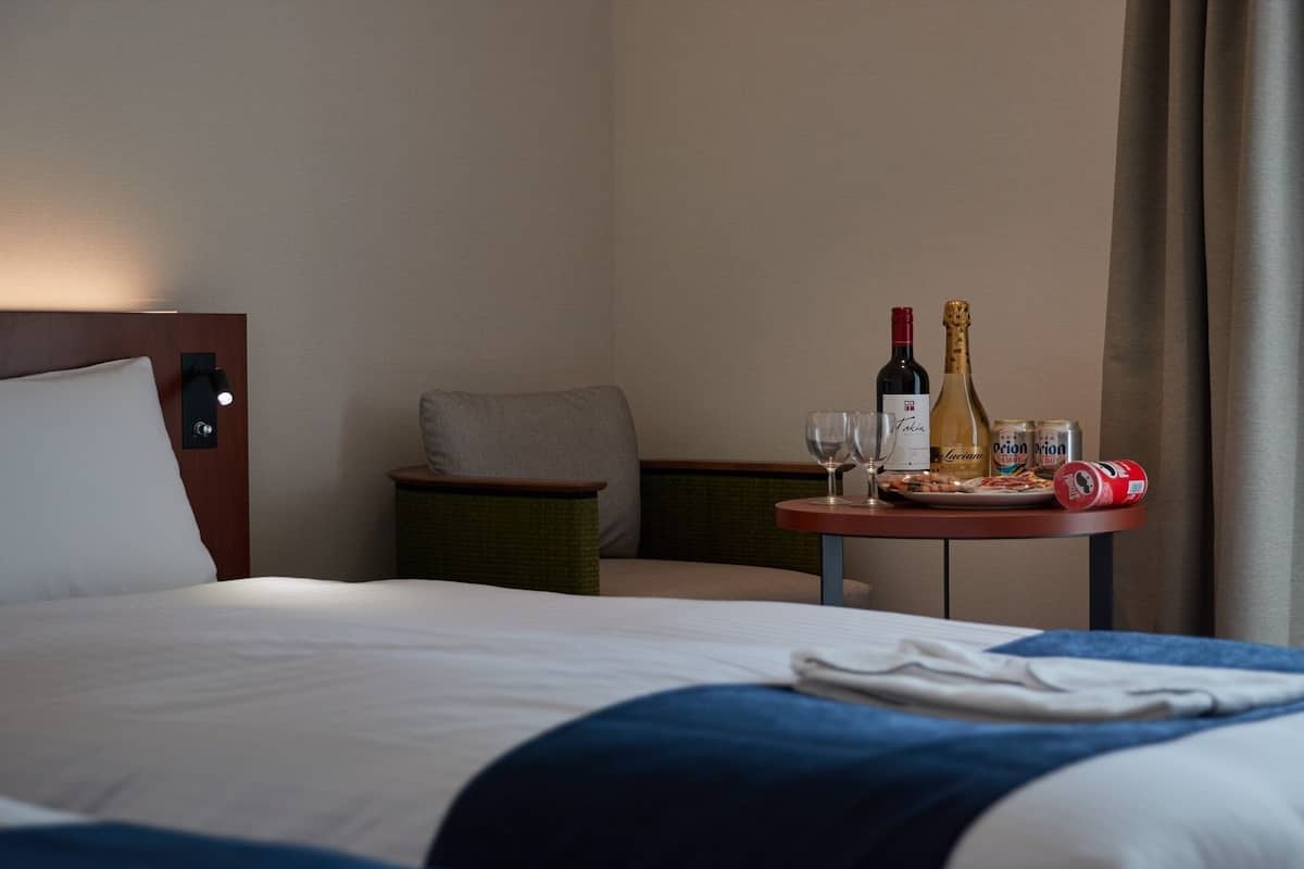 Quarto de hotel com cama bem arrumada, uma mesinha com garrafa de vinho, copos e uma pizza.